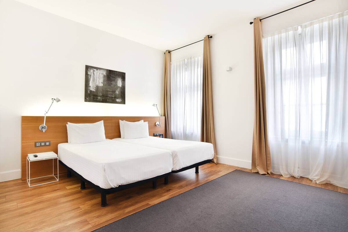 Quarto de hotel com cama de casal, mesa de cabeceira, abajur, quadro na parede e cortinas finas.