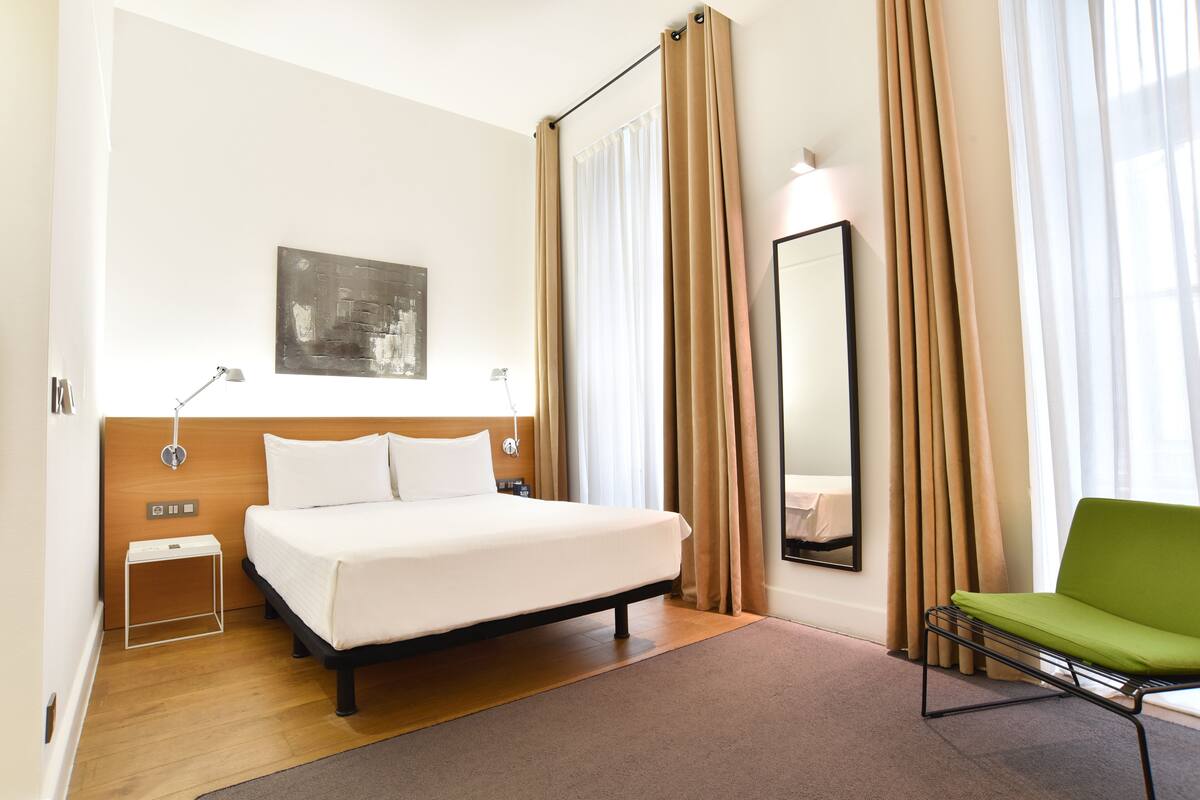 Quarto de hotel com cama, mesa de cabeceira, luminária, cadeira verde, espelho grande e um quadro na parede.