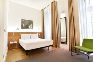 Quarto de hotel com cama, mesa de cabeceira, luminária, cadeira verde, espelho grande e um quadro na parede.