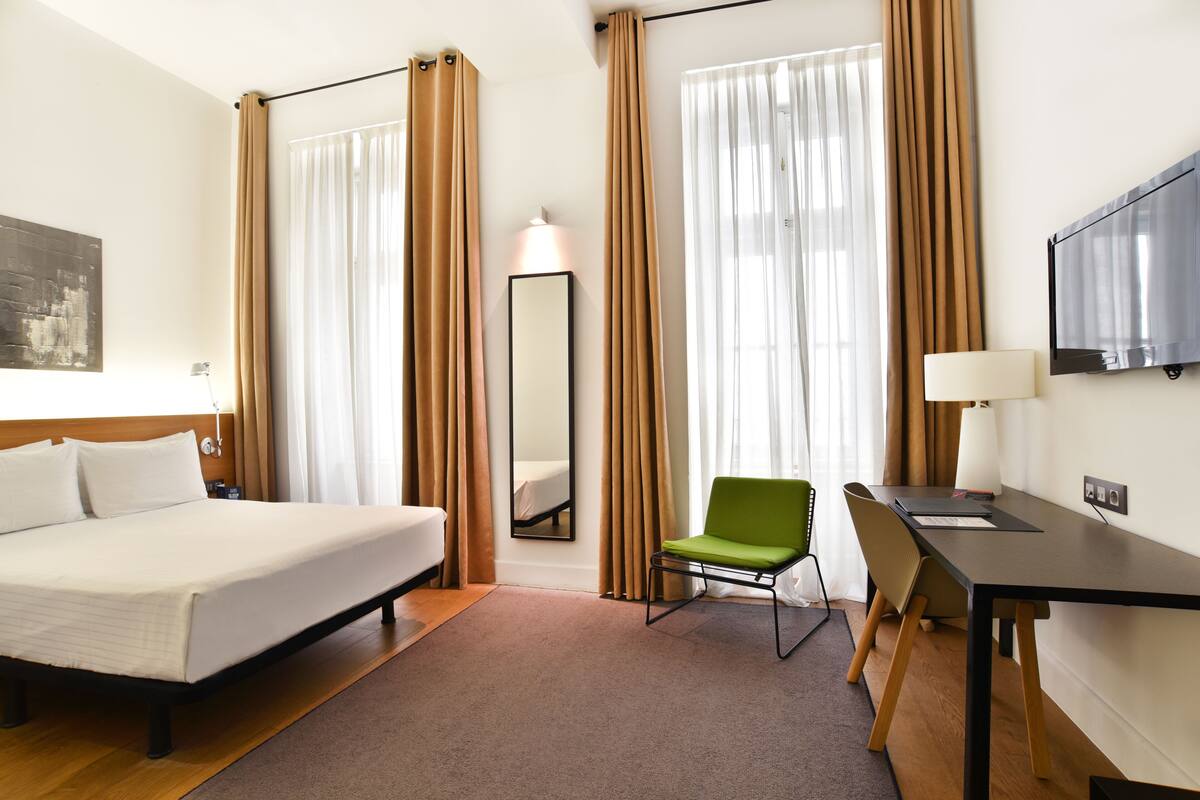 Quarto de hotel com cama, escrivaninha, cadeira, televisão e espelho.