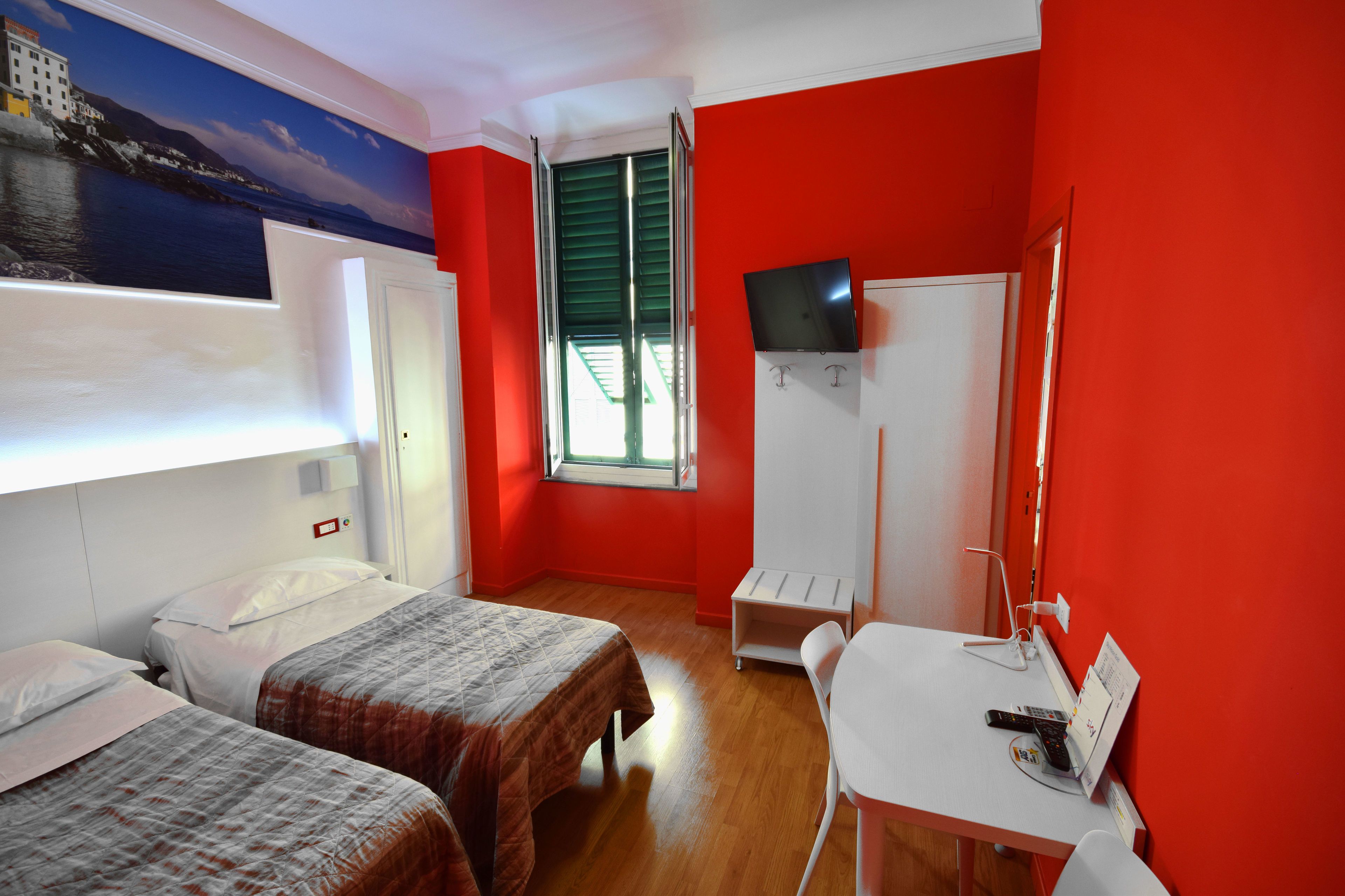 Quarto com duas camas, uma escrivaninha, uma televisão e uma janela com persianas verdes.