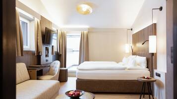 Quarto de hotel moderno com sofá, cama, pequena mesa com frutas, televisão e luminária.