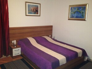Quarto com cabeceira de madeira, colcha listrada em roxo e branco, mesa de cabeceira com abajur e duas fotos emolduradas na parede.