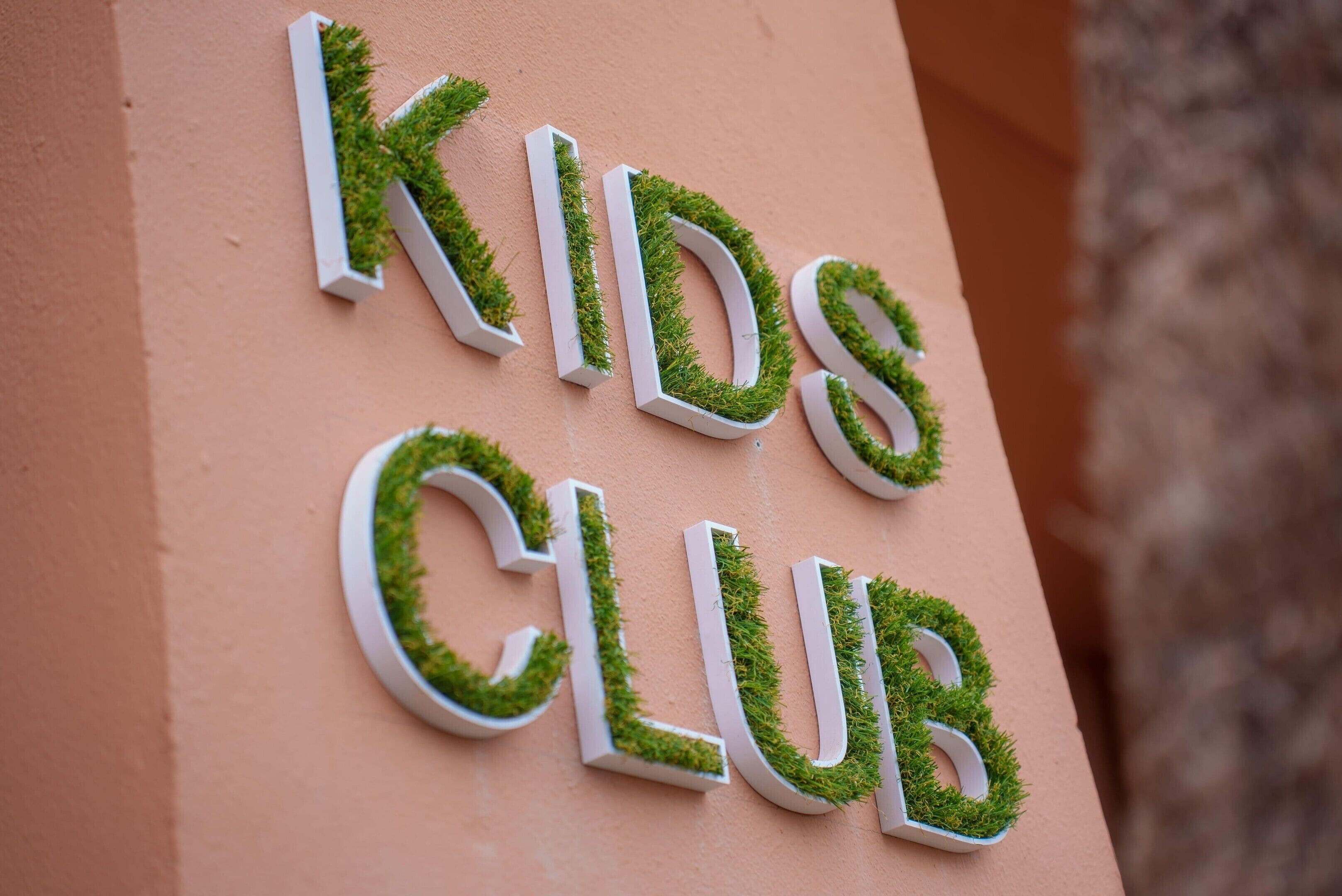 Um sinal com as palavras 'KIDS CLUB' em letras brancas, com bordas que lembram relva verde.