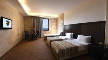 Quarto de hotel com duas camas, uma escrivaninha e uma cadeira. Há uma janela com cortinas, um quadro na parede e uma mesa de cabeceira com abajur.