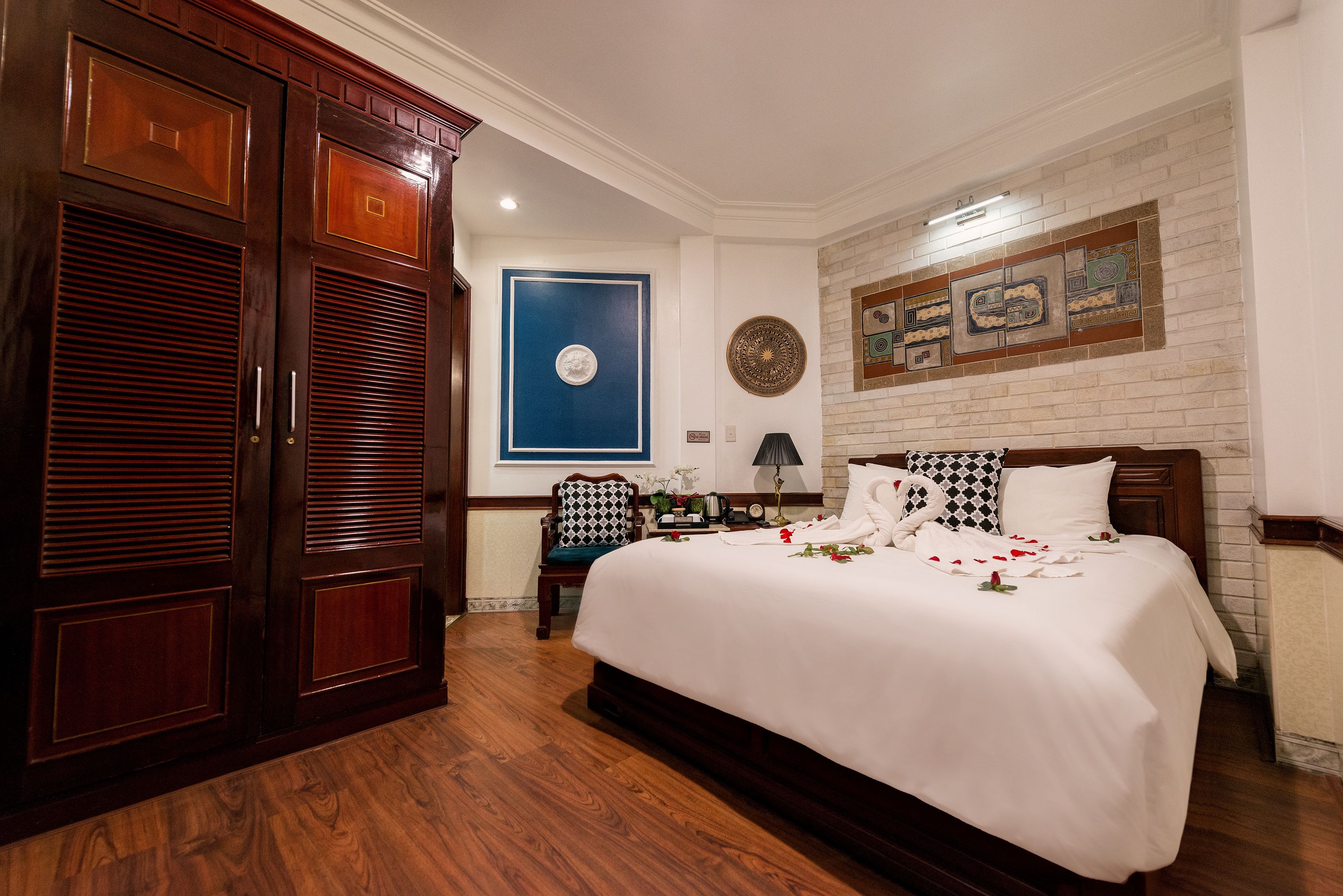 Quarto de hotel com um armário de madeira grande, uma cama com lençóis brancos e pétalas de rosa, uma parede azul de destaque e um objeto decorativo na parede.
