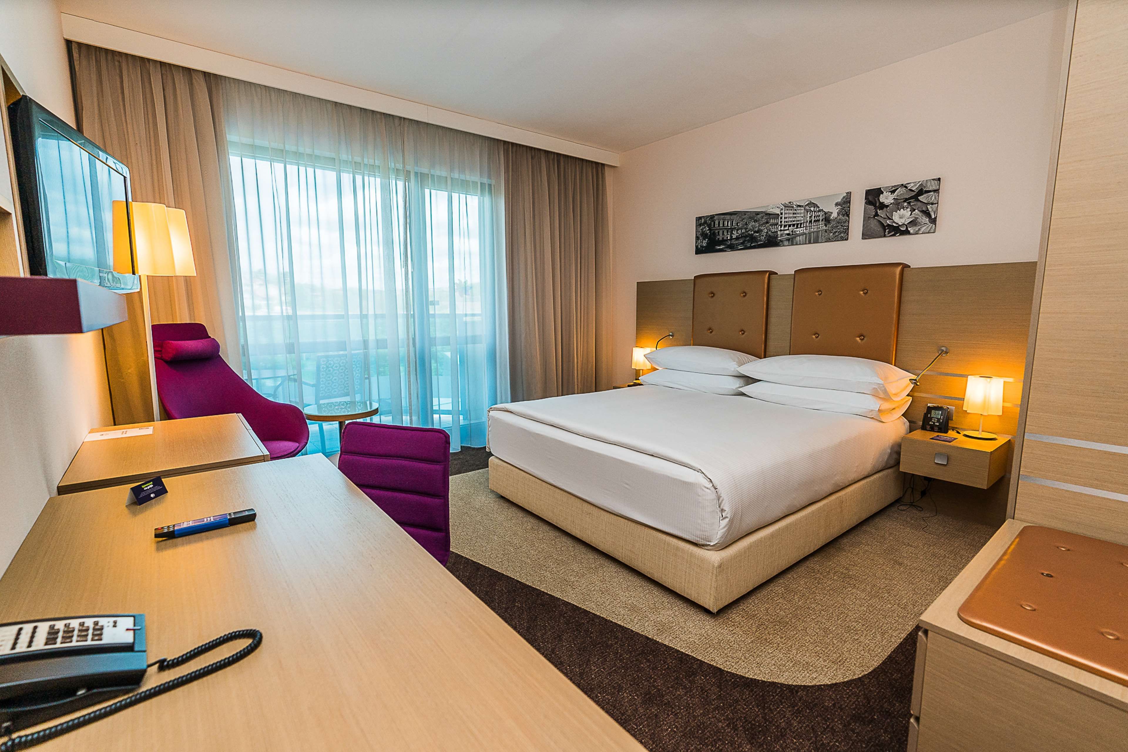 Quarto de hotel com cama, escrivaninha, cadeira, mesa de cabeceira e janela com cortinas.