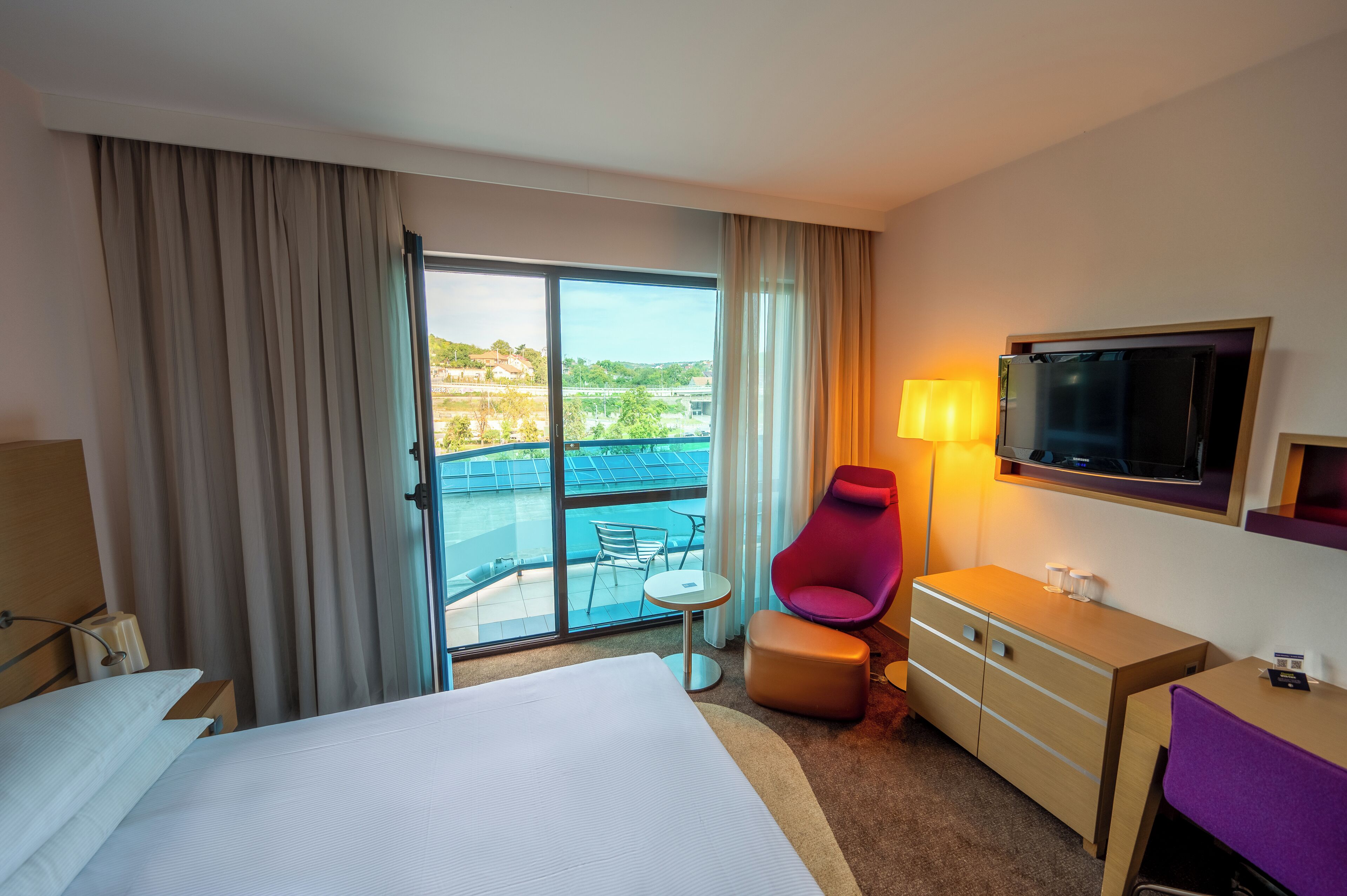 Quarto de hotel com cama, escrivaninha, cadeira, televisão e varanda com vista para a cidade.