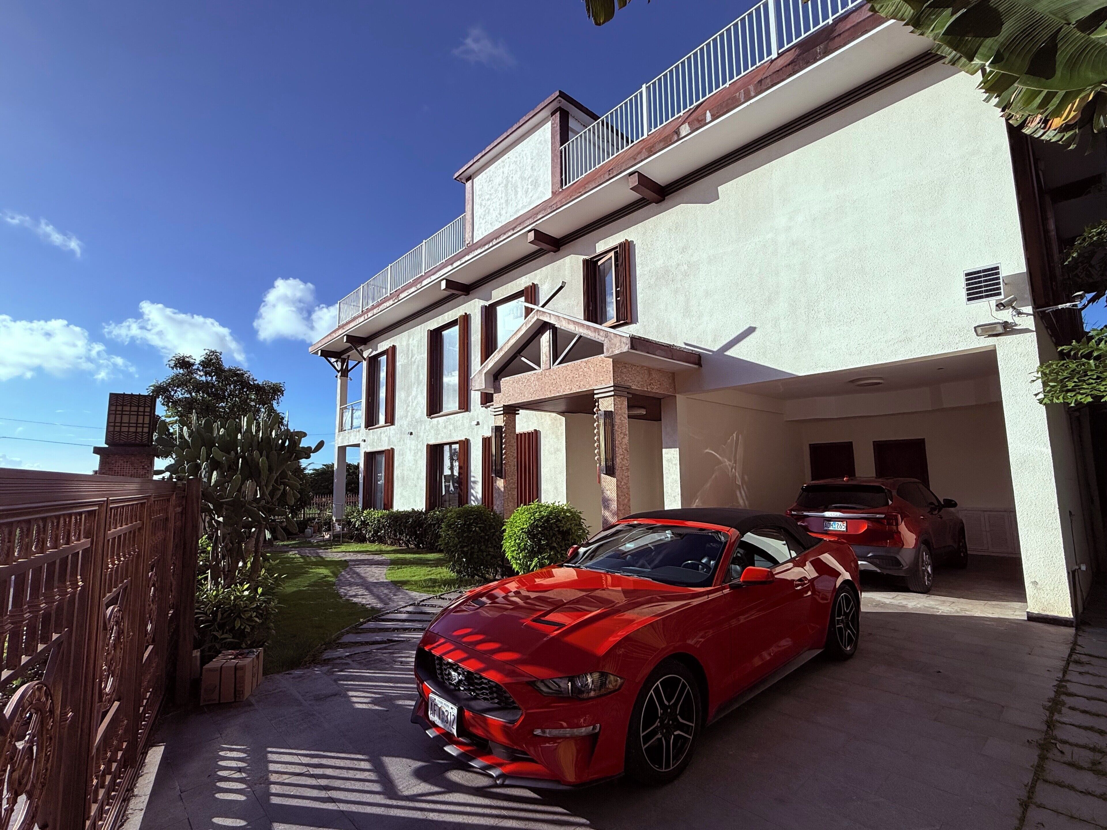 Uma casa de dois andares com garagem, um carro esportivo vermelho estacionado na entrada e um jardim bem cuidado.