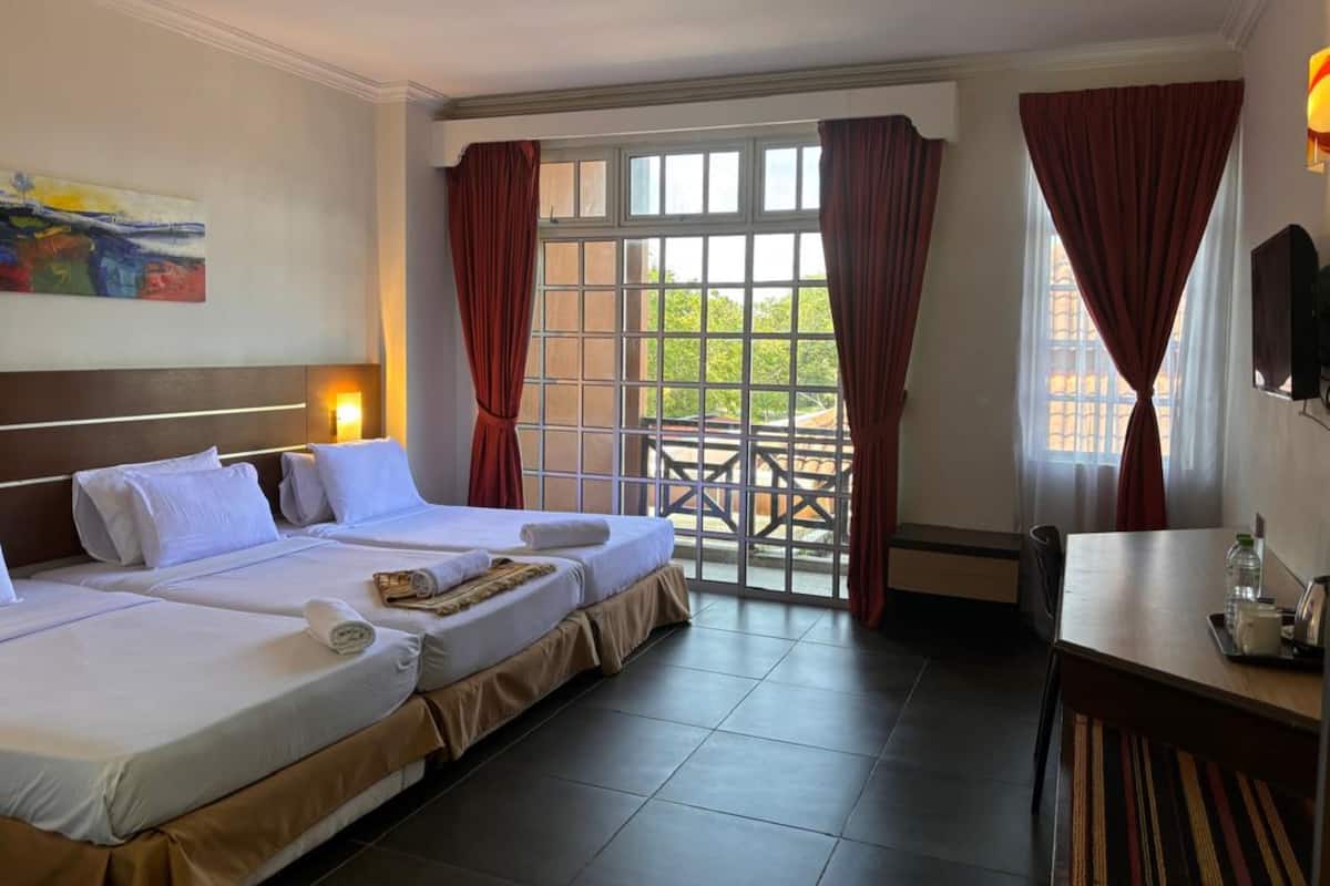 Quarto de hotel com duas camas, uma escrivaninha, uma cadeira e uma janela grande com cortinas.