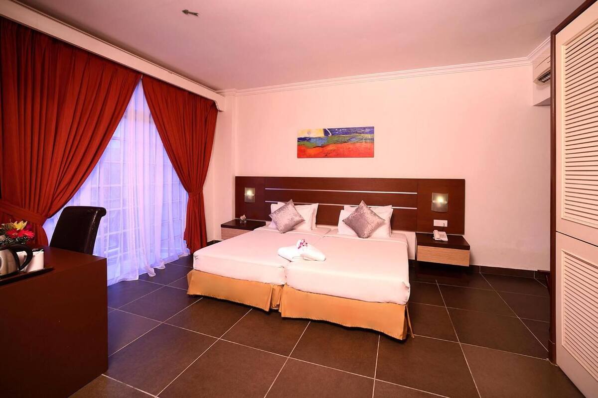 Quarto de hotel com uma cama grande, uma escrivaninha, uma cadeira, um quadro na parede e uma janela com cortinas vermelhas.