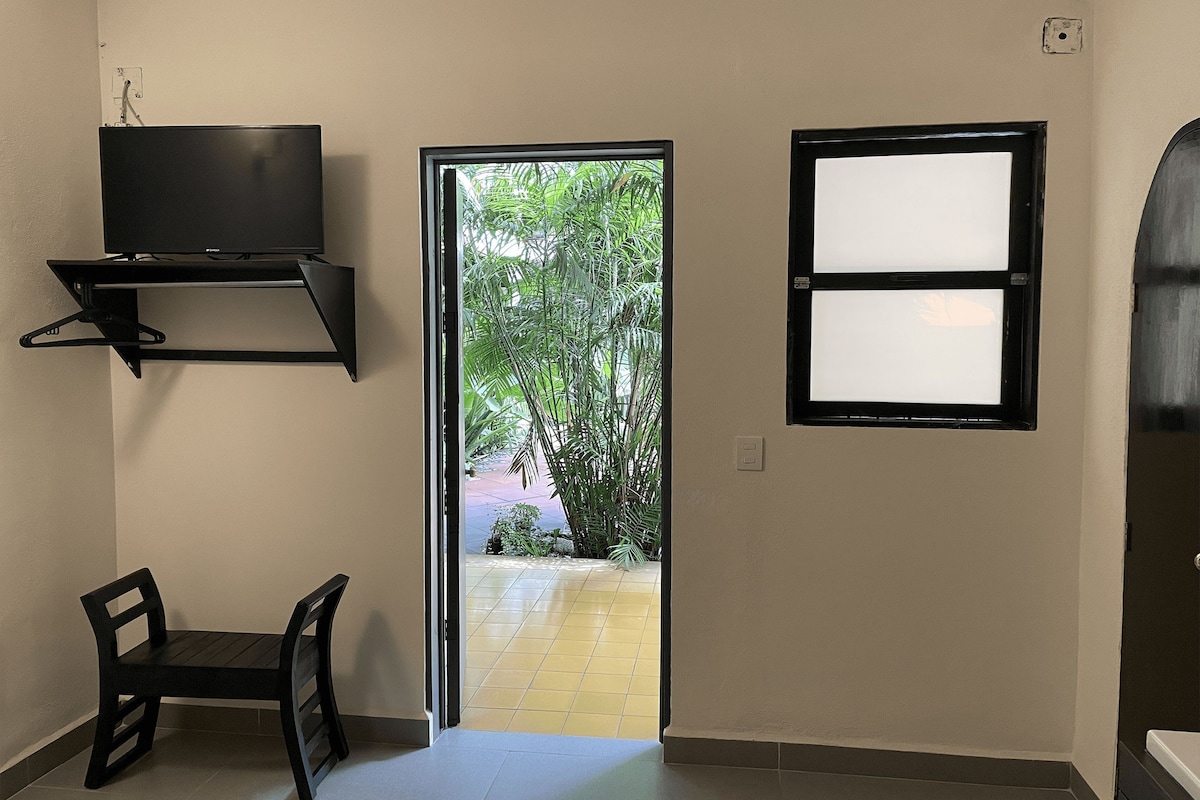 Quarto com televisão sobre suporte, porta que dá para um jardim e janela com persianas.