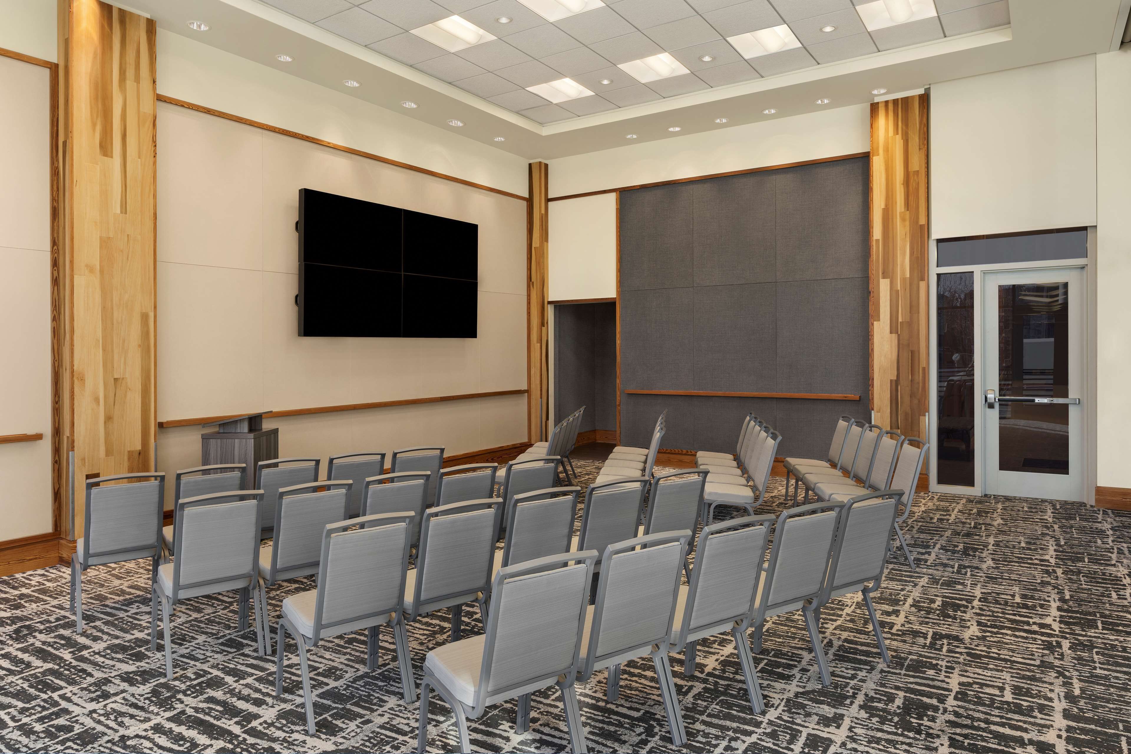 Uma sala de conferência com fileiras de cadeiras, uma tela grande e um tapete estampado.