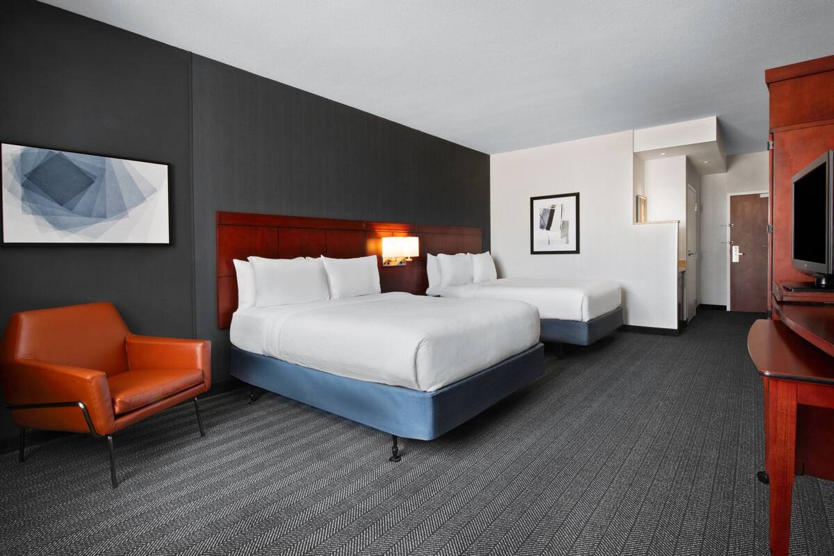Quarto de hotel com uma cama grande, uma cama menor, um sofá, uma televisão e obras de arte nas paredes.