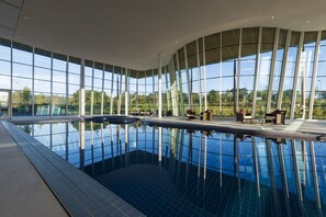 Uma grande piscina interior com paredes de vidro transparente, rodeada por áreas de estar e vista para o exterior.