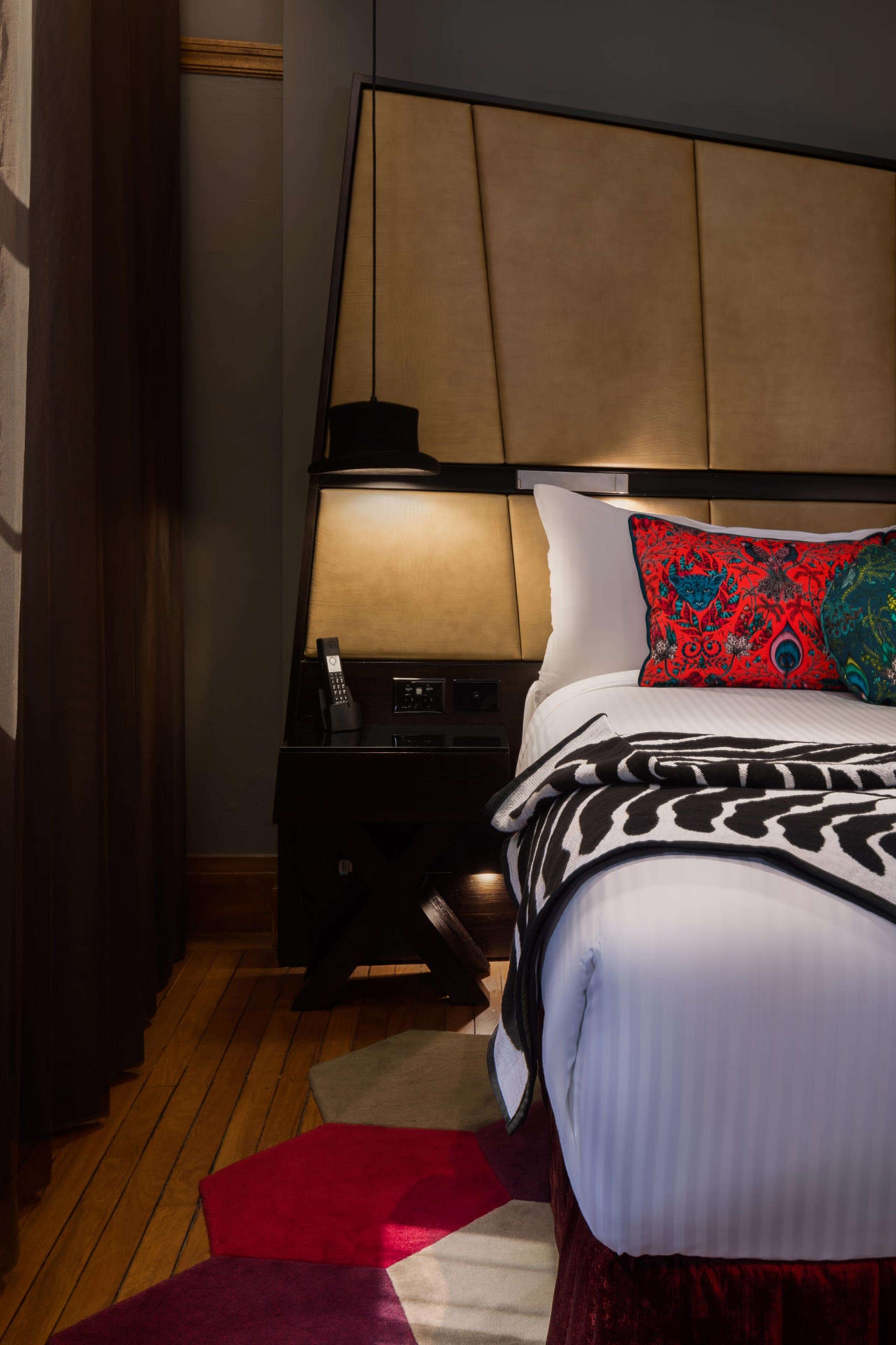 Quarto de hotel com uma cama, uma mesa de cabeceira com abajur e um tapete vermelho e branco.