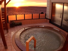 Uma banheira de hidromassagem com vista para o pôr do sol sobre o oceano, rodeada por piso de azulejos e guarda-corpos de madeira.