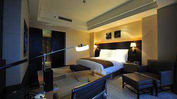 Quarto de hotel com cama, duas mesas de cabeceira, escrivaninha com cadeira, sofá e divisória de vidro.