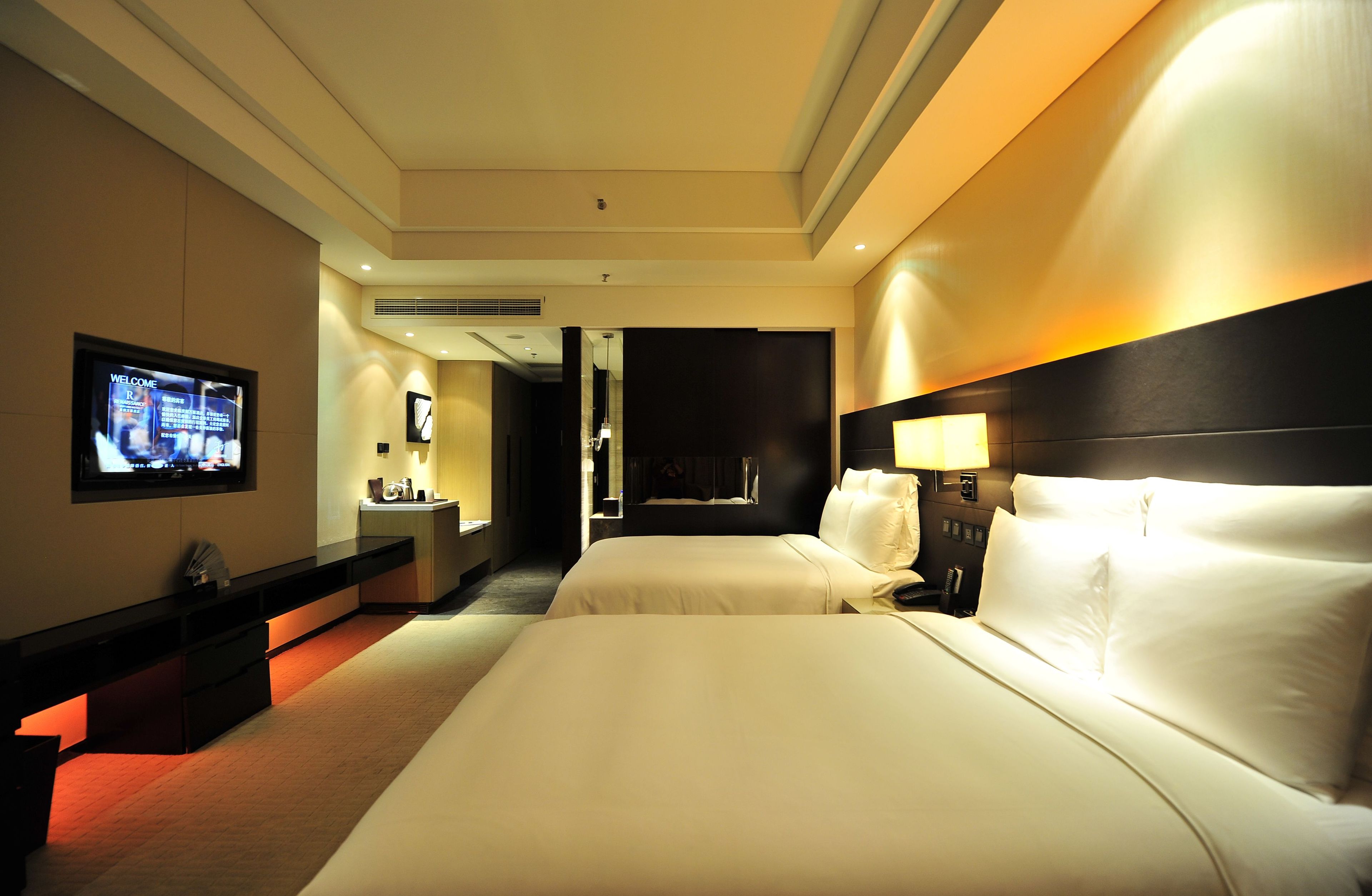 Quarto de hotel com uma cama grande, televisão na parede, mesa de cabeceira e criado-mudo.