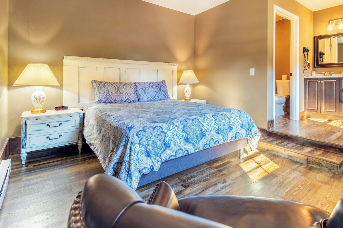 Quarto com cama, abajures, mesa de cabeceira, cômoda e banheiro visível por uma porta aberta.