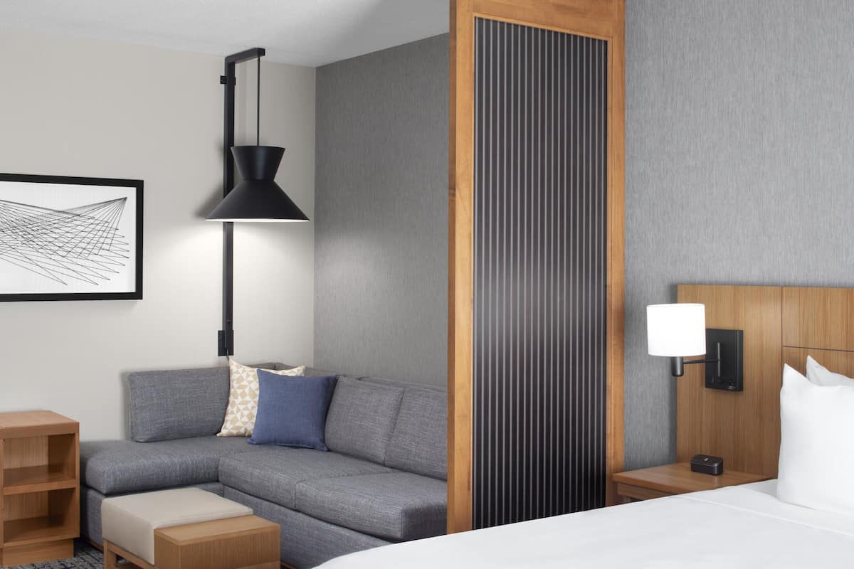 Quarto de hotel moderno com sofá cinzento, mesa de cabeceira de madeira, mesa lateral, luminária e uma obra de arte emoldurada na parede.