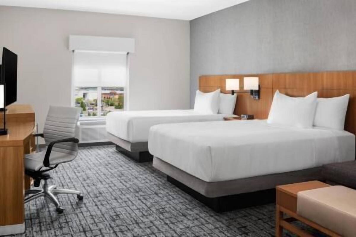 Quarto de hotel com duas camas, uma escrivaninha, uma cadeira e uma janela com vista.
