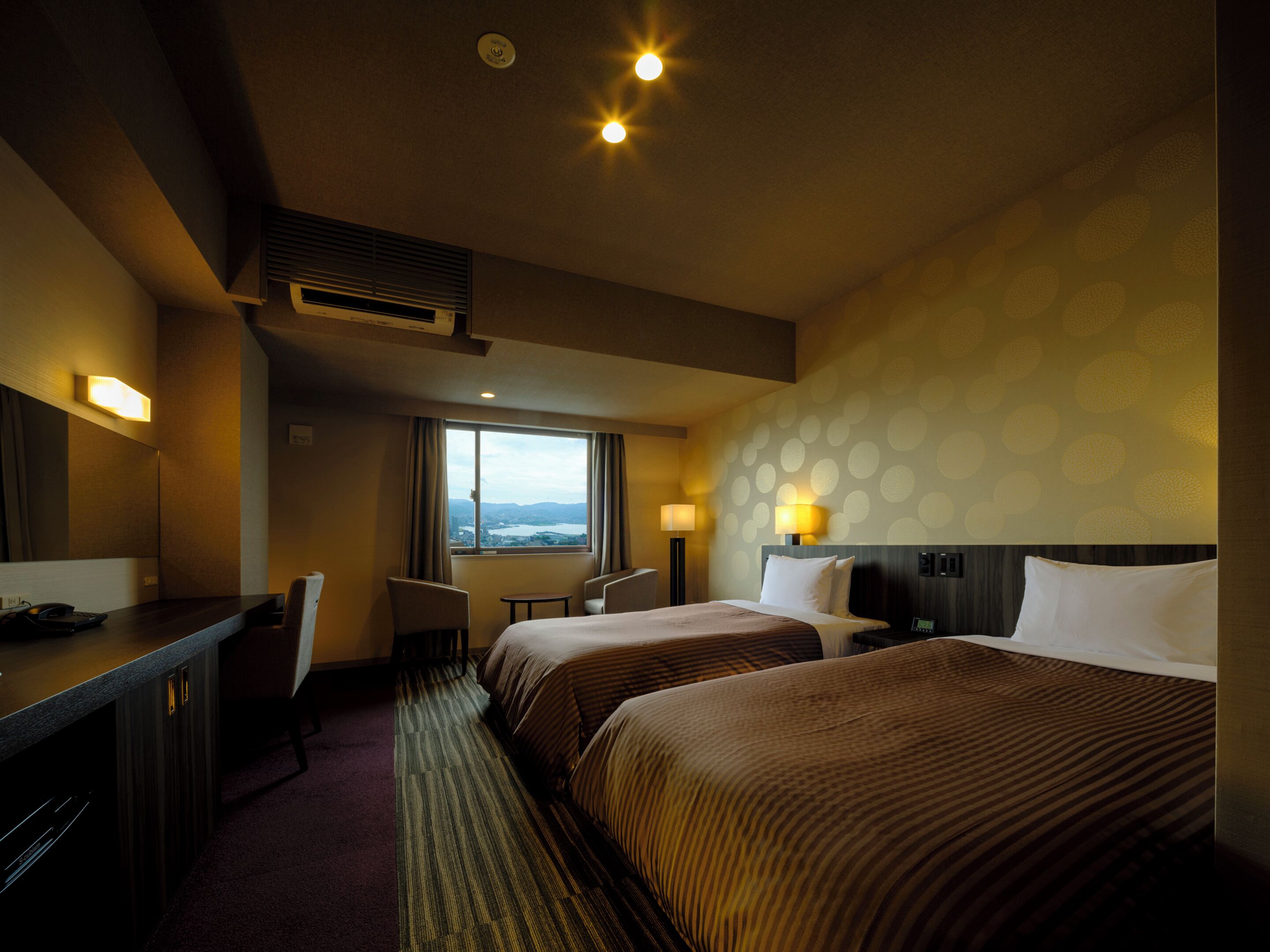 Quarto de hotel com duas camas, uma escrivaninha, uma cadeira e janela com vista.