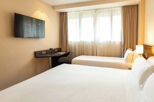 Quarto de hotel com duas camas, uma escrivaninha, uma cadeira, uma televisão de tela plana e uma janela com cortinas.