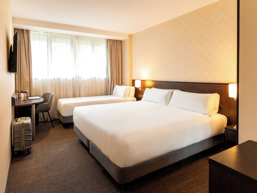 Quarto de hotel com duas camas, uma escrivaninha, uma cadeira, uma televisão e uma janela com cortinas.