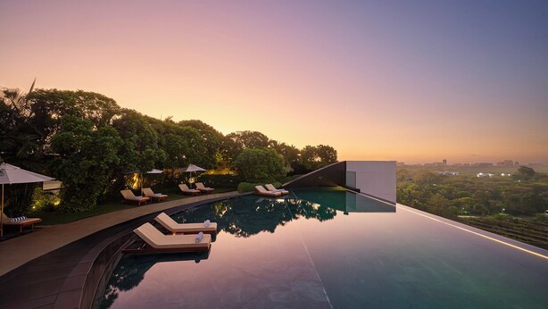 Uma piscina infinita com espreguiçadeiras e guarda-sóis, cercada por vegetação exuberante e um horizonte urbano ao fundo.