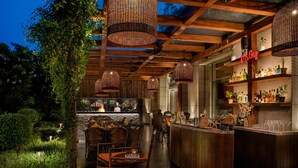 Uma área de bar com mobiliário de madeira, luminárias de vime suspensas e prateleiras com garrafas e plantas.