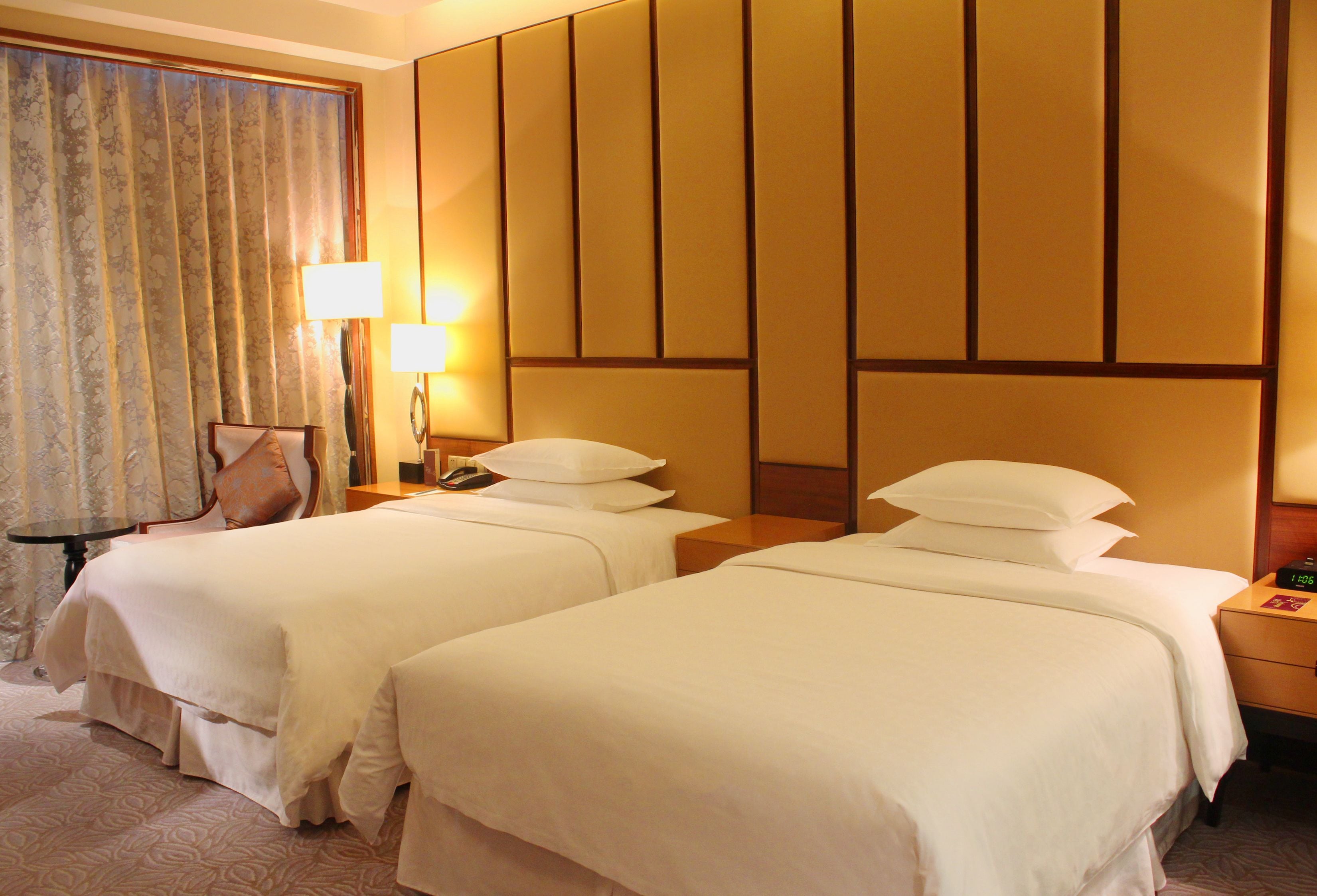 Quarto de hotel com duas camas, uma escrivaninha, uma lâmpada e um relógio.
