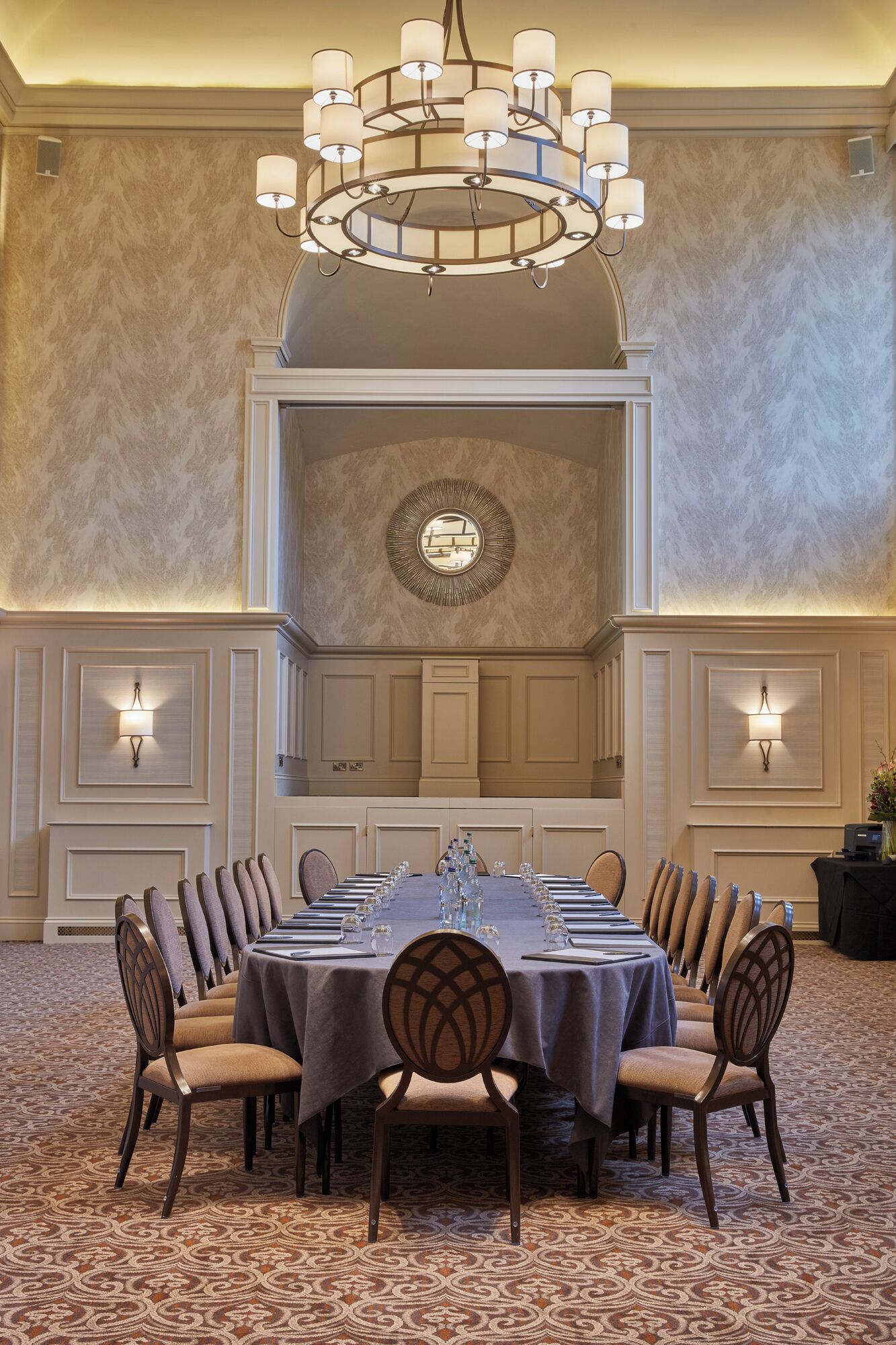 Uma sala de jantar formal com uma grande mesa redonda, várias cadeiras, um candelabro e arandelas nas paredes.