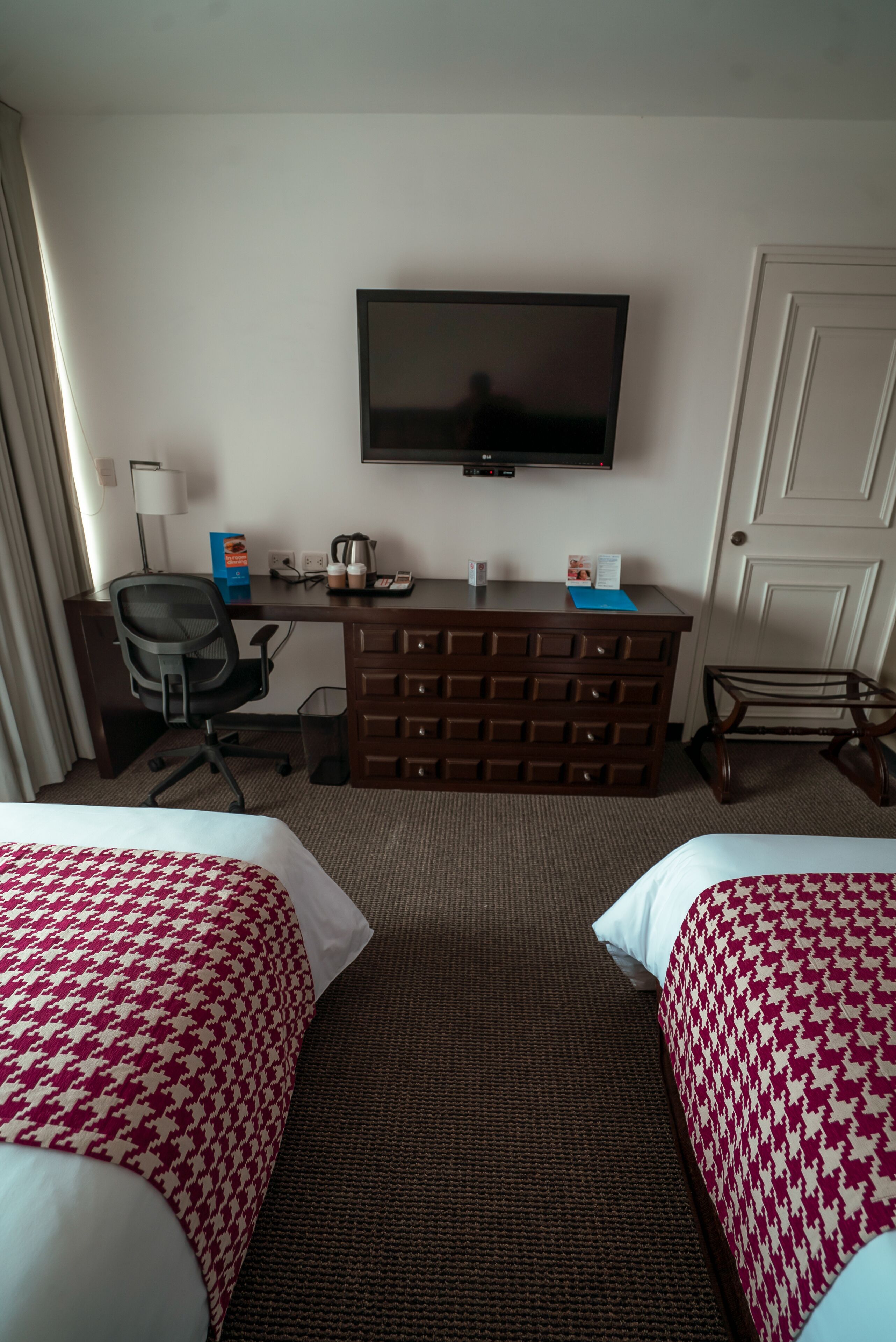 Quarto de hotel com duas camas, uma escrivaninha com cadeira, televisão, minibar e uma porta.