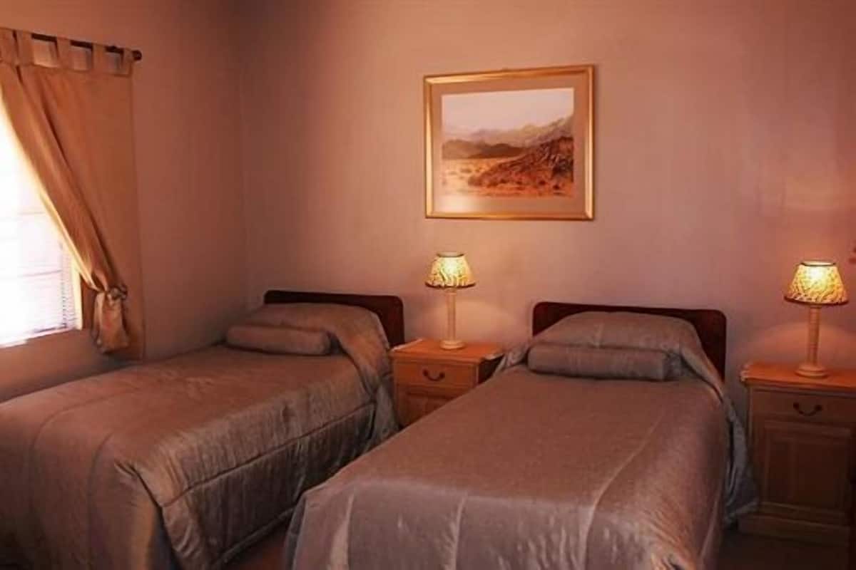 Quarto com duas camas de solteiro, cada uma com mesa de cabeceira e luminária. Há uma janela com cortinas e um quadro pendurado na parede.