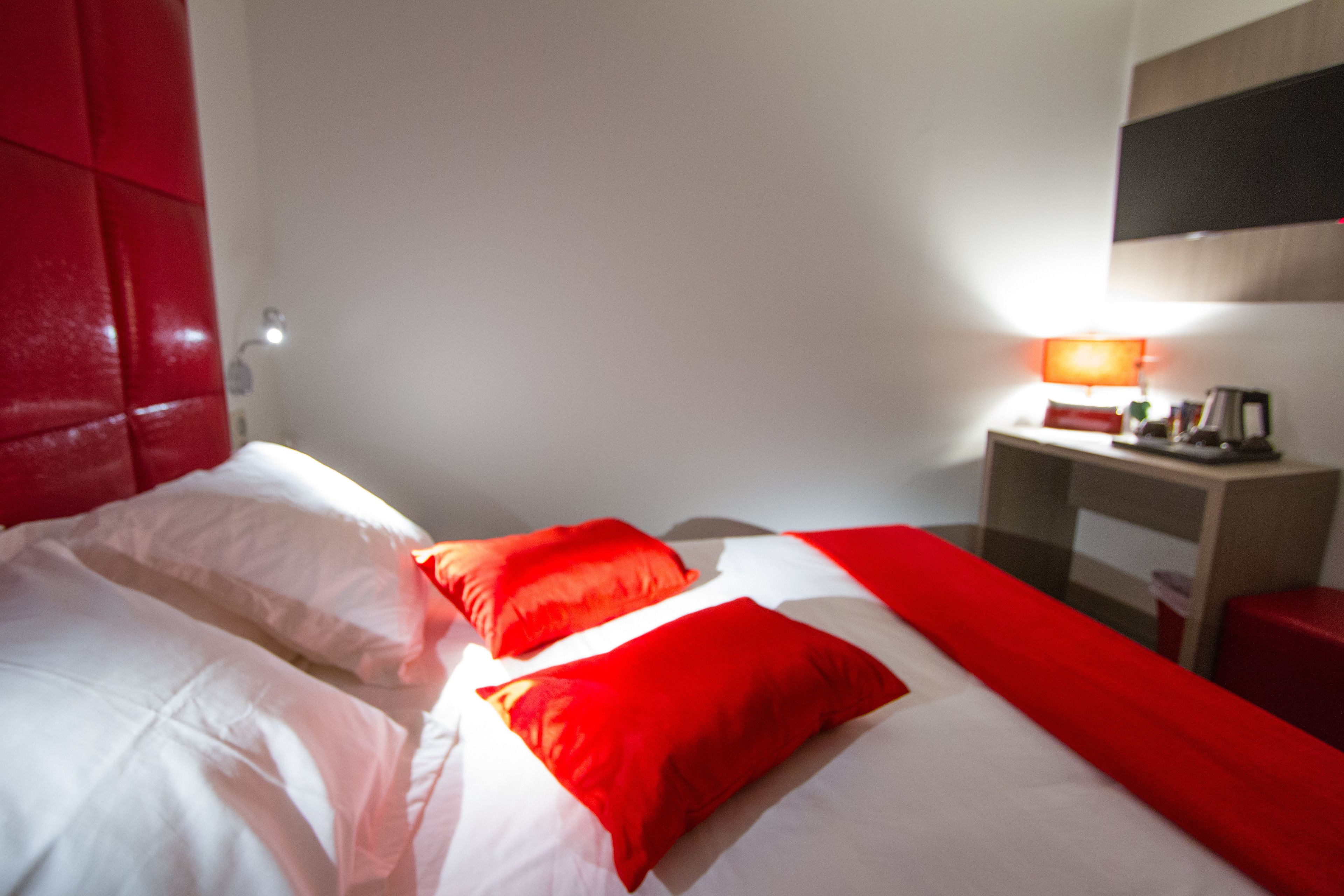 Quarto de hotel com cama, almofadas vermelhas, roupa de cama branca, mesa de cabeceira com abajur e televisão na parede.