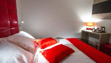 Quarto de hotel com cama, almofadas vermelhas, roupa de cama branca, mesa de cabeceira com abajur e televisão na parede.