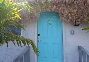 Uma porta turquesa com uma placa indicando o número 25, um telhado de palha e uma pequena luminária fixada na parede.