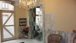 Um saguão de hotel com um candelabro, uma mesa de recepção, uma cadeira estampada, uma planta em vaso com flores e um mapa na parede.