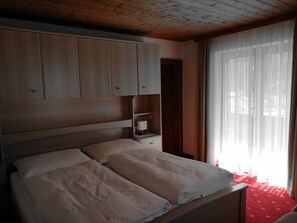 Quarto com cabeceira de madeira, duas camas com roupa de cama branca, mesa de cabeceira e janela com cortinas leves.