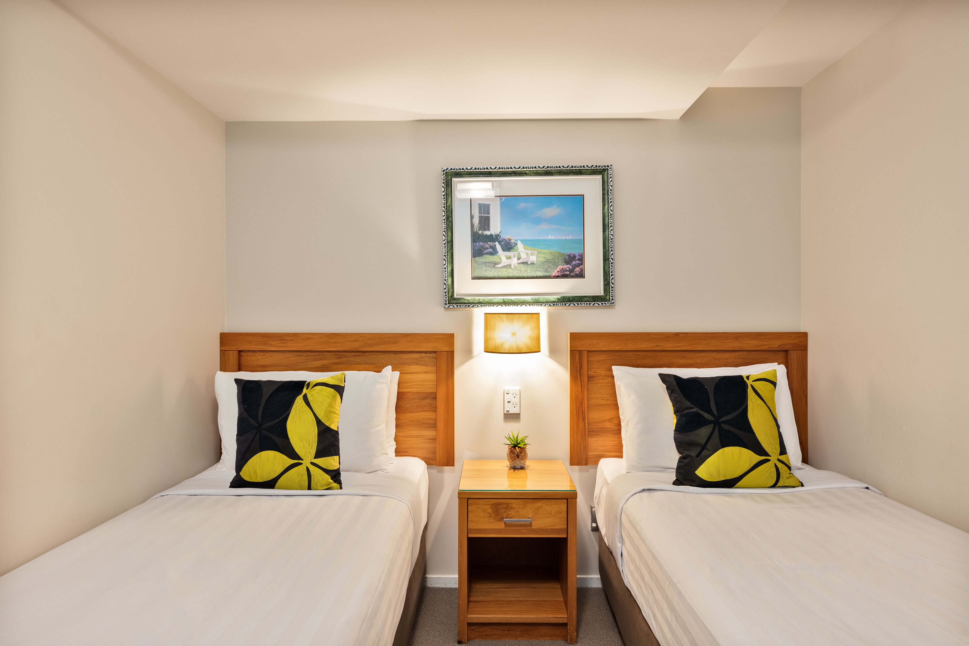 Quarto de hotel com duas camas, uma mesa de cabeceira com abajur e um quadro pendurado na parede.