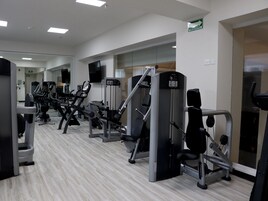Uma academia com diversos equipamentos de exercício, uma televisão de tela plana e uma placa indicando uma rota de evacuação.