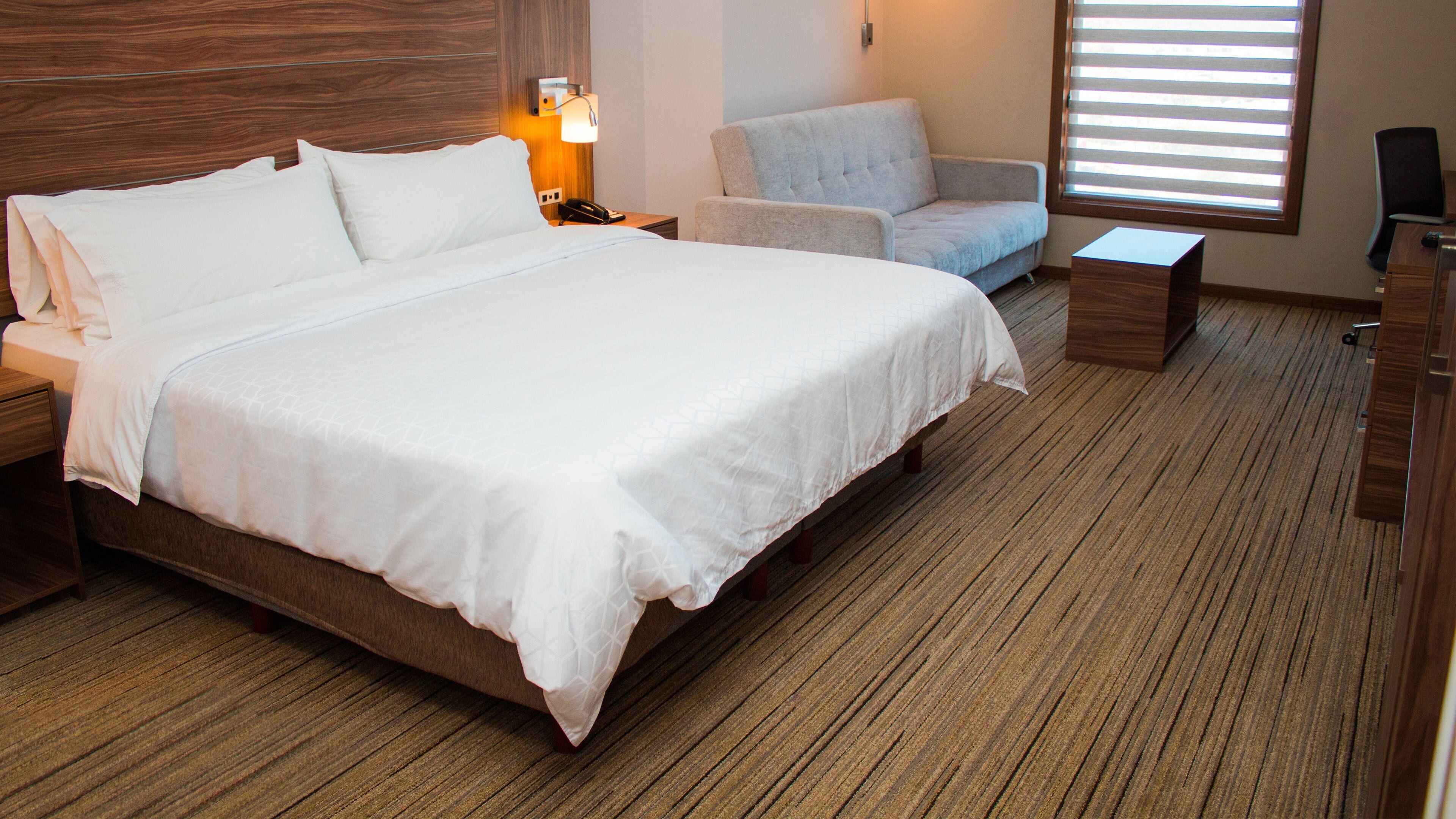 Quarto de hotel com uma cama grande, mesa de cabeceira, cadeira, pequena mesa e janela com persianas.