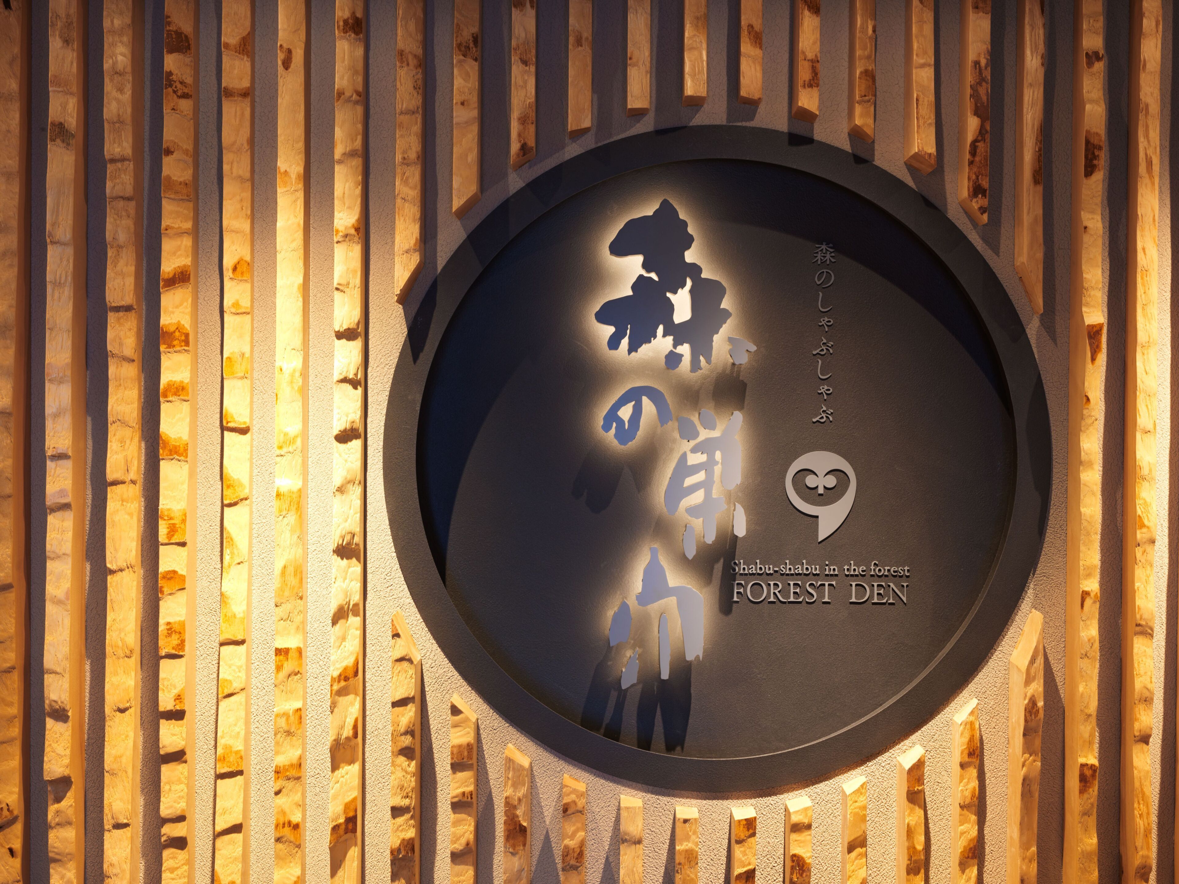Um sinal circular com caracteres japoneses e o silhueta de uma família, rodeada por ripas verticais de madeira.