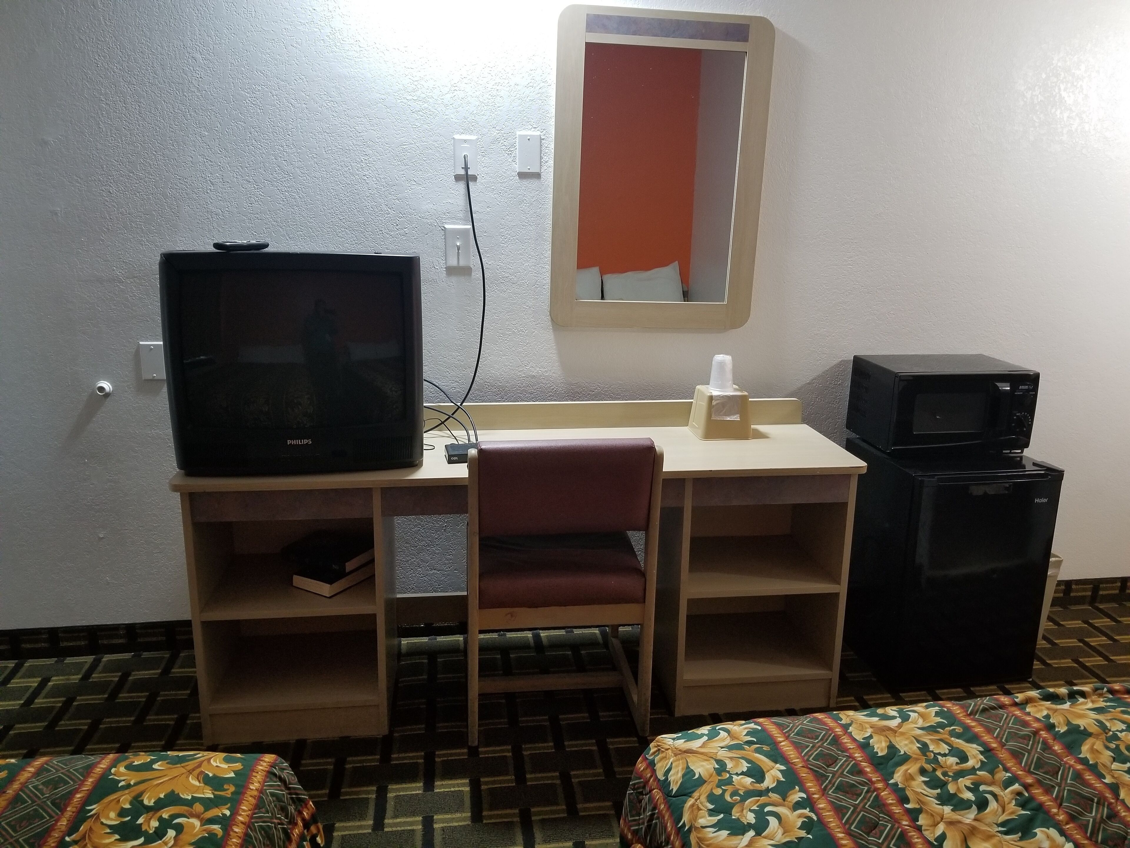 Quarto de hotel com escrivaninha de madeira, televisão, micro-ondas, espelho e duas camas com roupa de cama estampada.