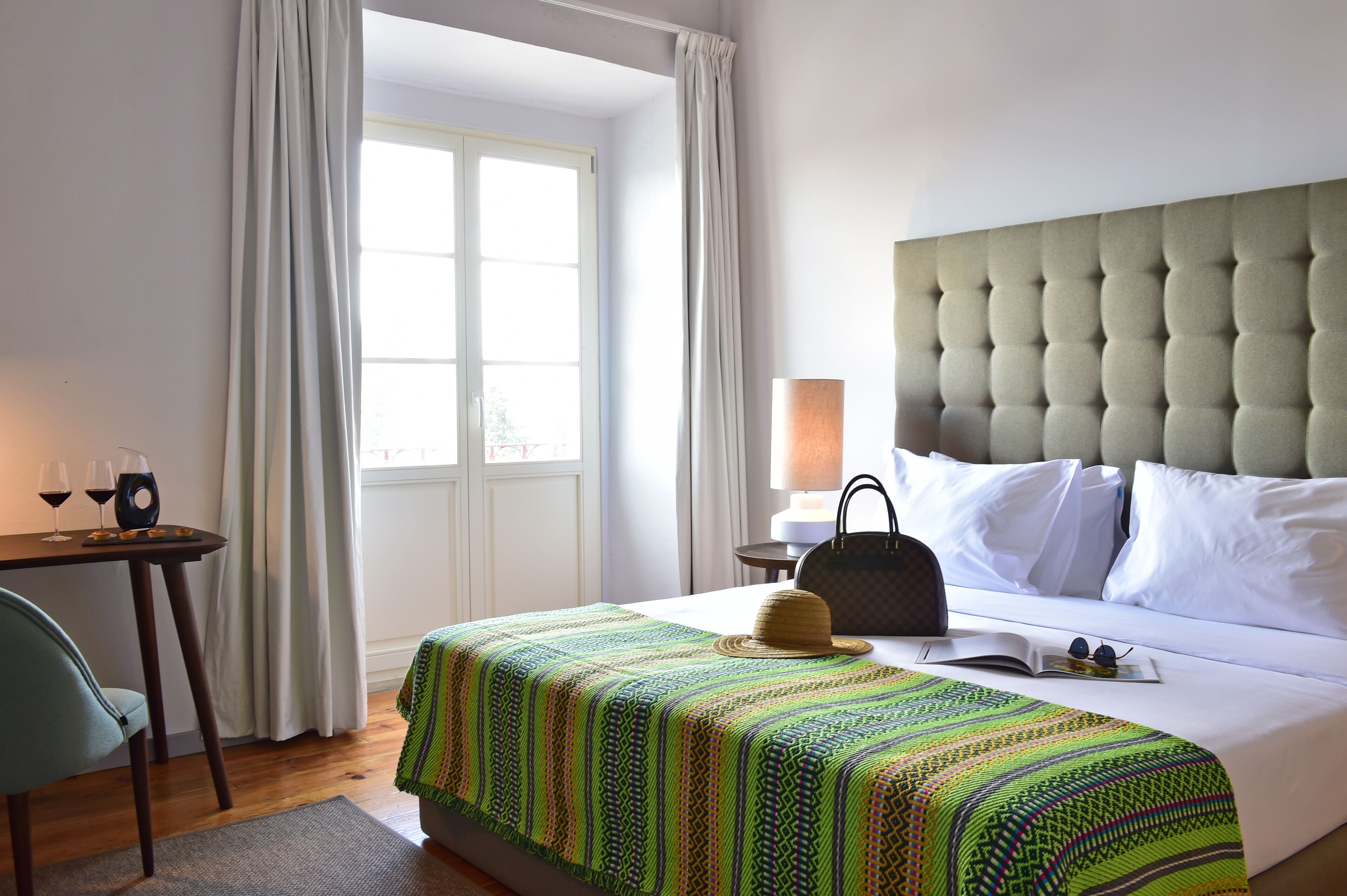 Quarto de hotel com uma cama, mesa de cabeceira com abajur, chapéu e um livro.
