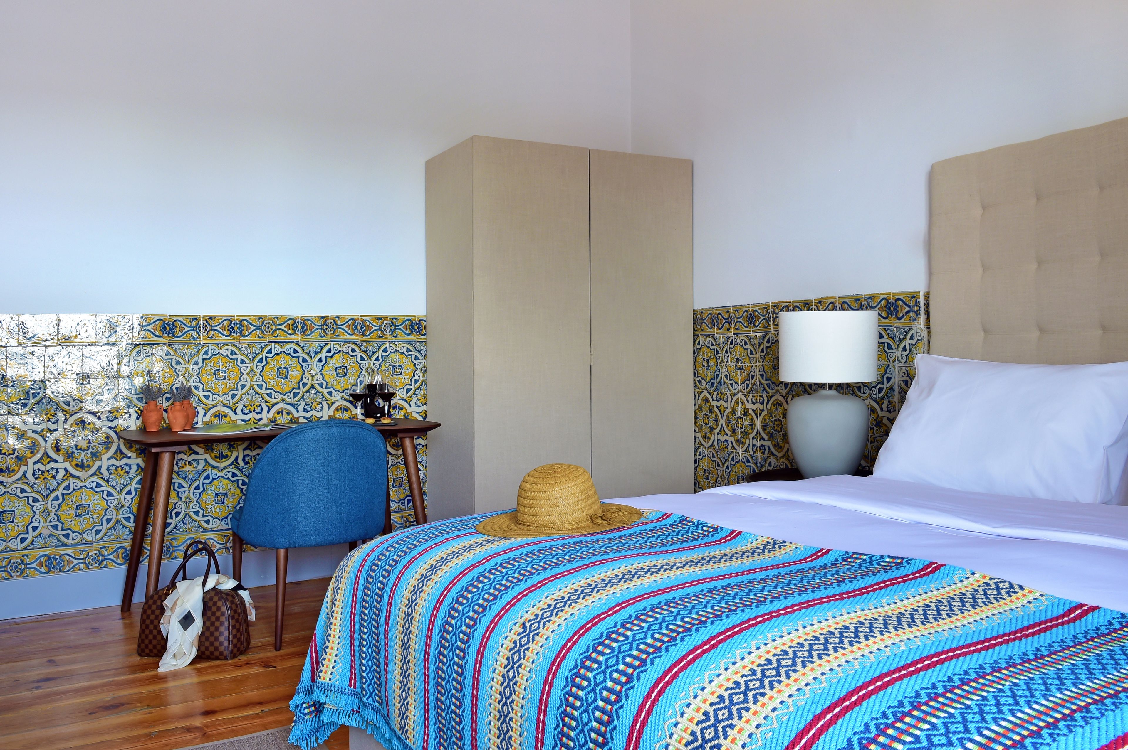 Quarto de hotel com uma cama, uma escrivaninha, uma cadeira, uma luminária e uma mala.