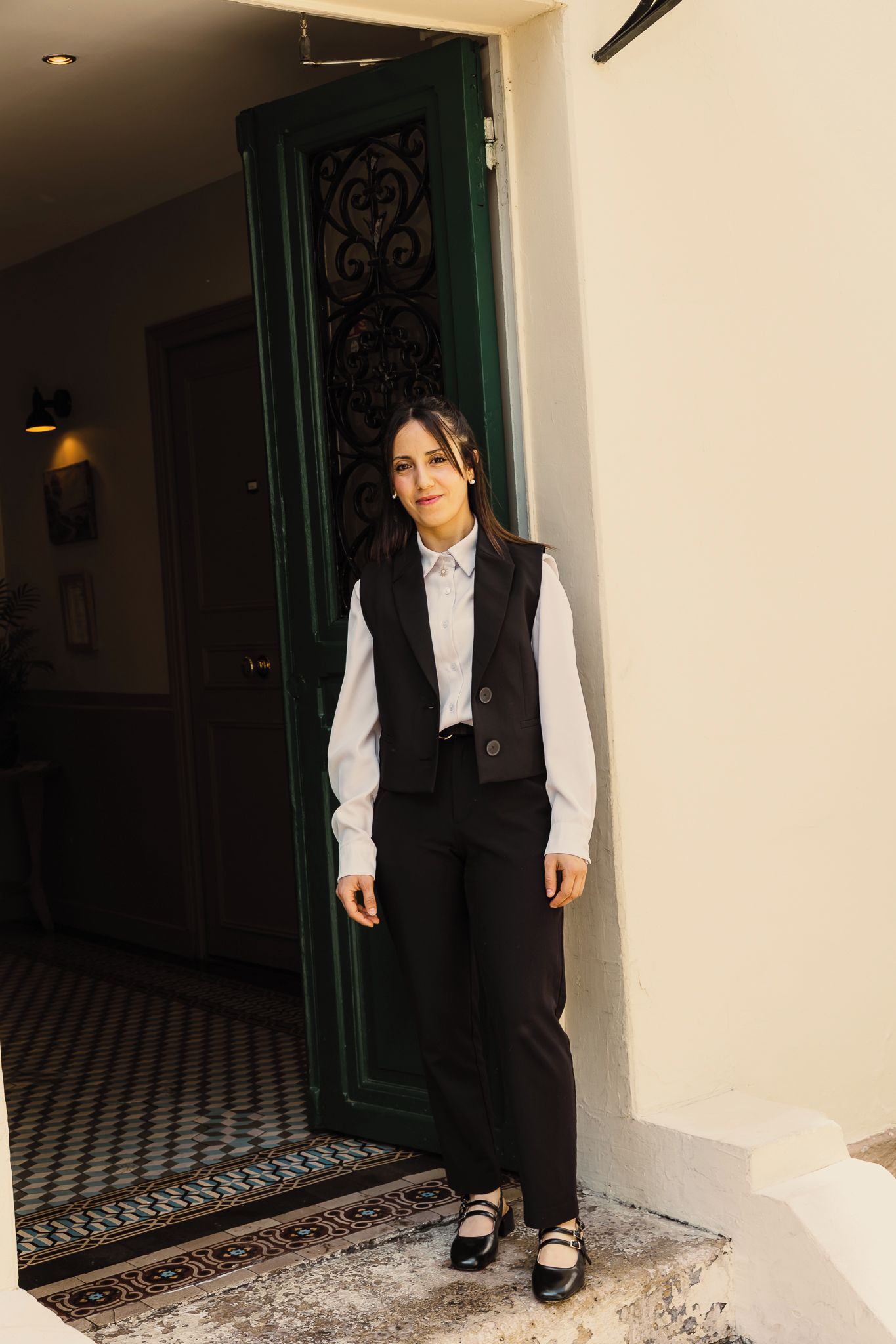 Uma pessoa em frente à entrada de um edifício, vestindo camisa branca, colete preto e calças pretas.