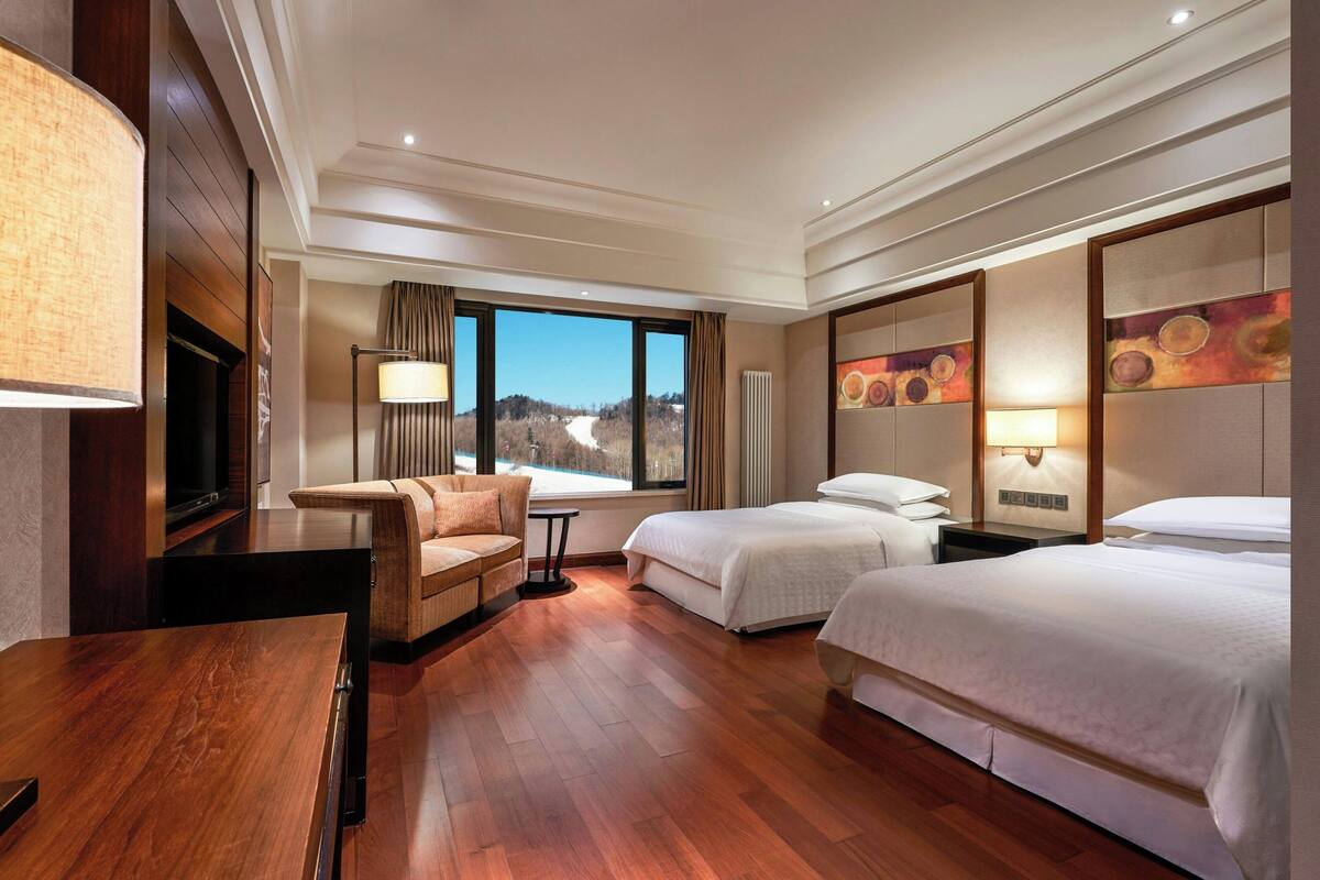 Quarto de hotel com piso de madeira, duas camas, um sofá, uma TV e janela com vista para árvores.