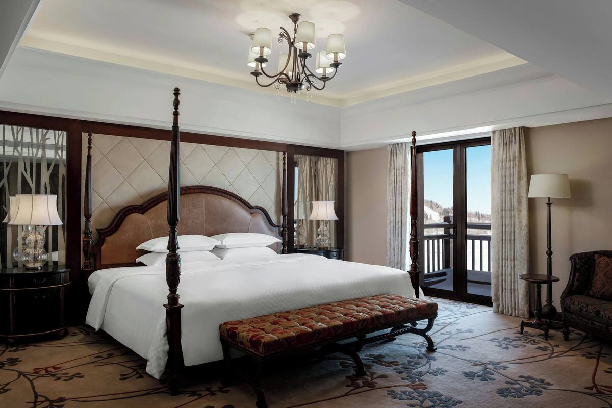 Uma cama com dossel, um banco, um candelabro e vista para o exterior através de uma varanda.